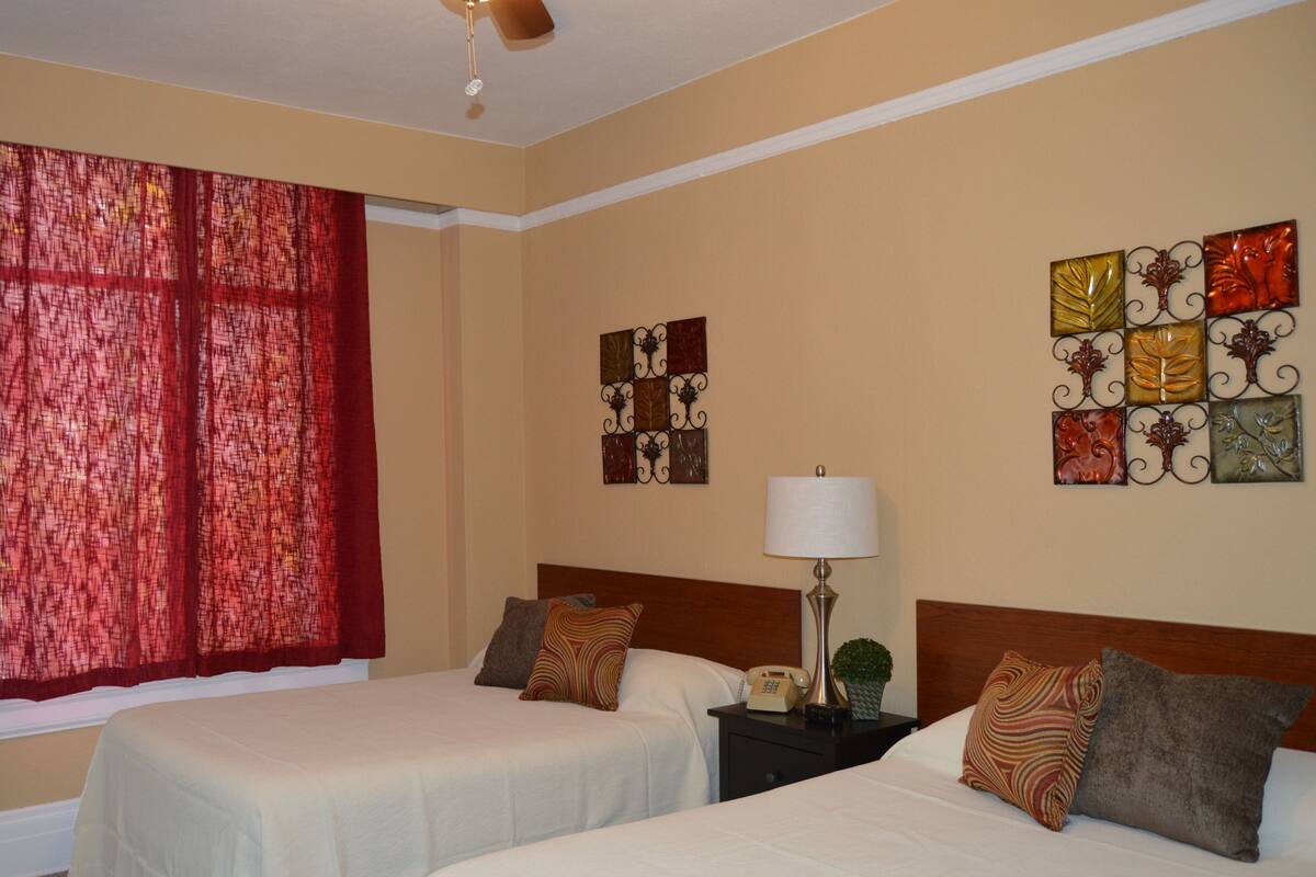 Quarto de hotel com duas camas, janela com cortinas vermelhas, mesa de cabeceira com abajur e uma obra de arte na parede.