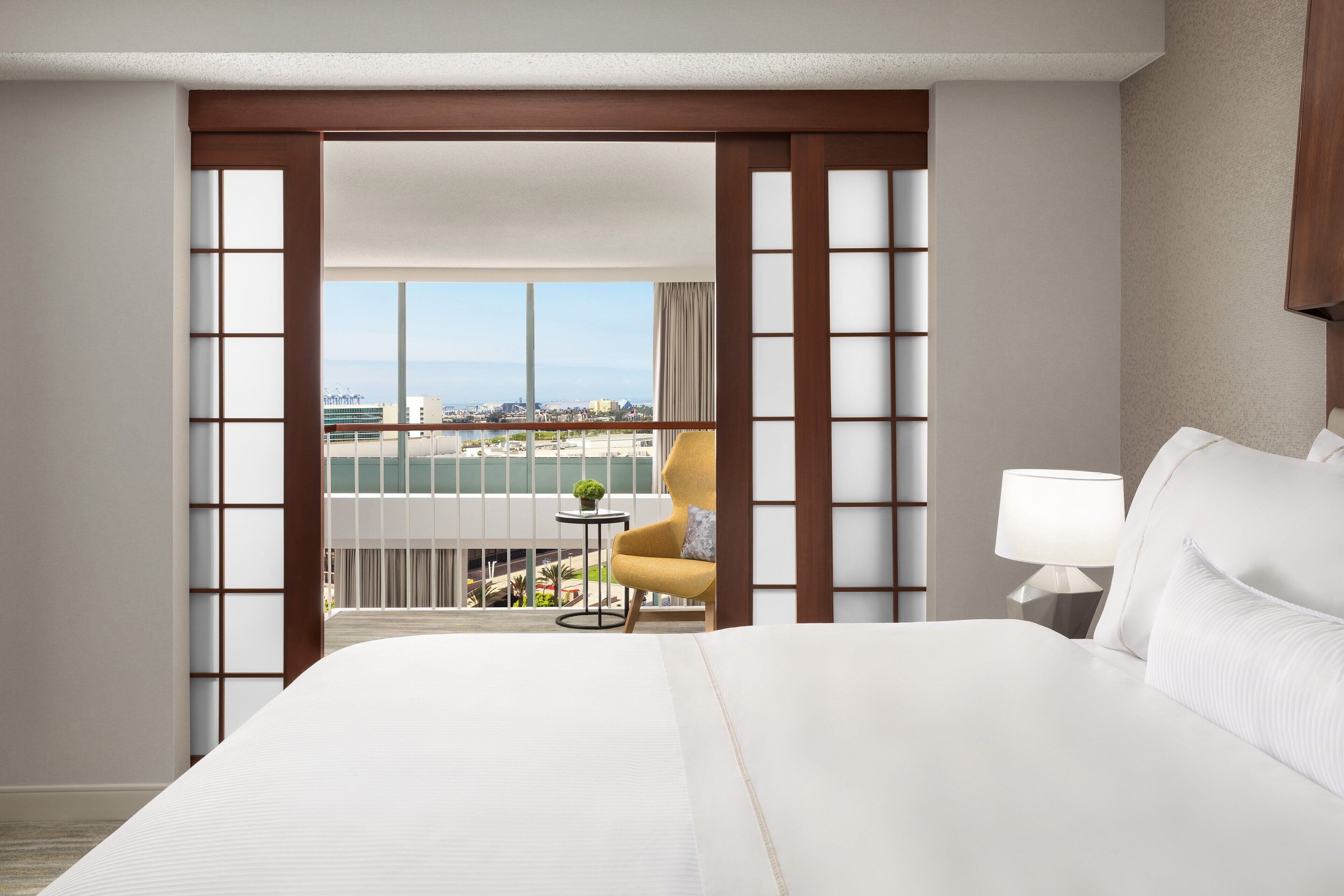 Quarto de hotel com uma cama grande, uma mesa pequena com abajur, uma cadeira e vista da cidade através de uma porta deslizante.