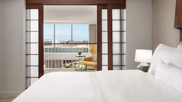 Quarto de hotel com uma cama grande, uma mesa pequena com abajur, uma cadeira e vista da cidade através de uma porta deslizante.