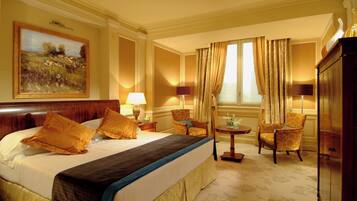 Quarto de hotel com uma cama grande, duas poltronas, uma mesa pequena e um quadro na parede.