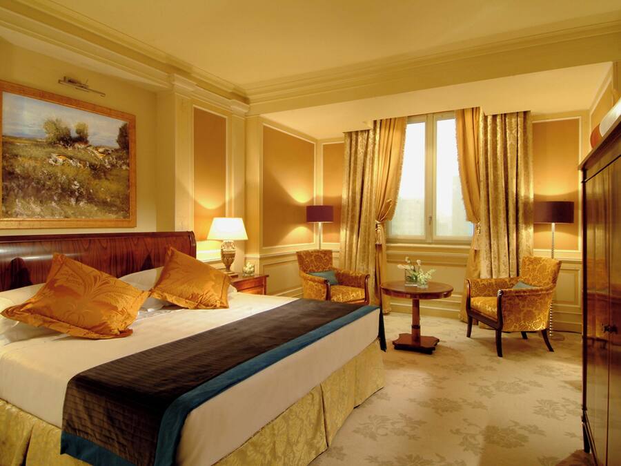 Quarto de hotel com uma cama grande, duas poltronas, uma mesa pequena e um quadro na parede.