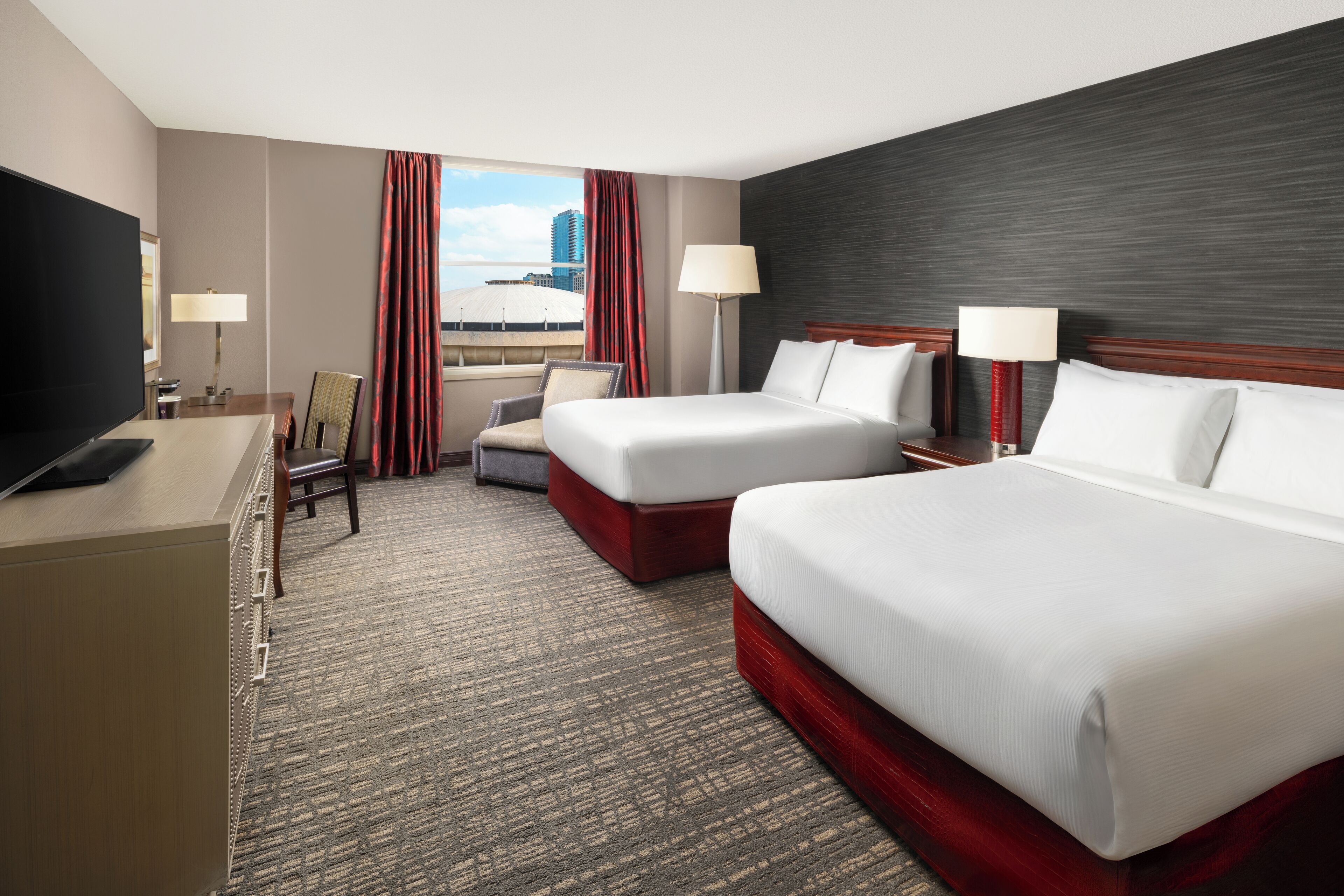 Quarto de hotel com duas camas, uma escrivaninha, televisão e vista para a paisagem urbana.