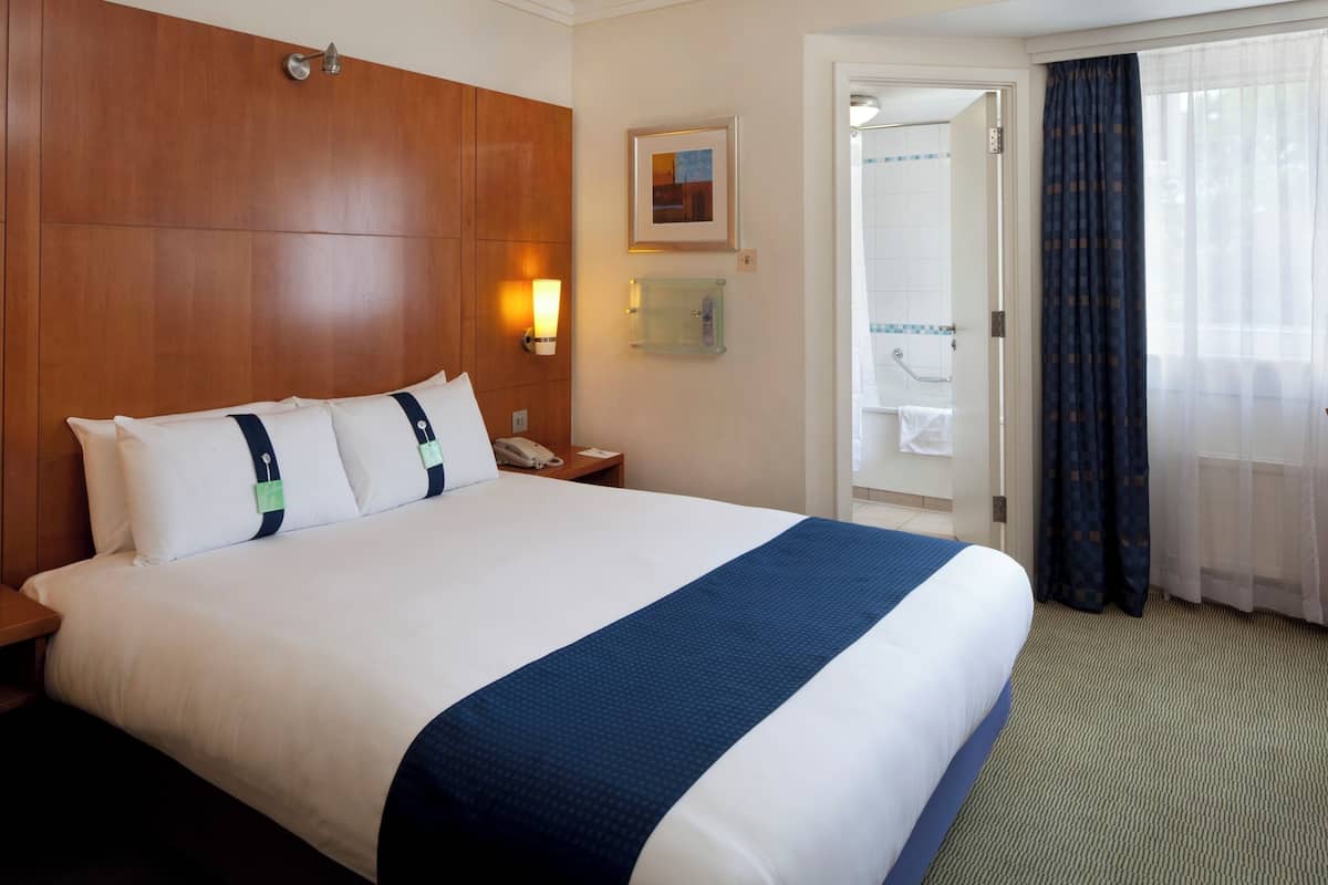 Quarto de hotel com cama grande, cabeceira de madeira, mesa de cabeceira com abajur, prateleira de vidro com artigos de higiene pessoal e porta para o banheiro.