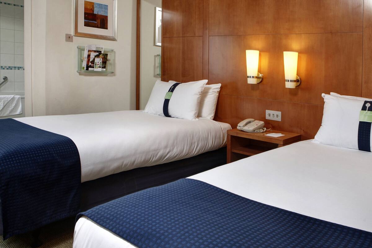 Quarto de hotel com duas camas, cabeceira de madeira, luminárias fixadas na parede, telefone ao lado da cama e uma pequena estante com folhetos.