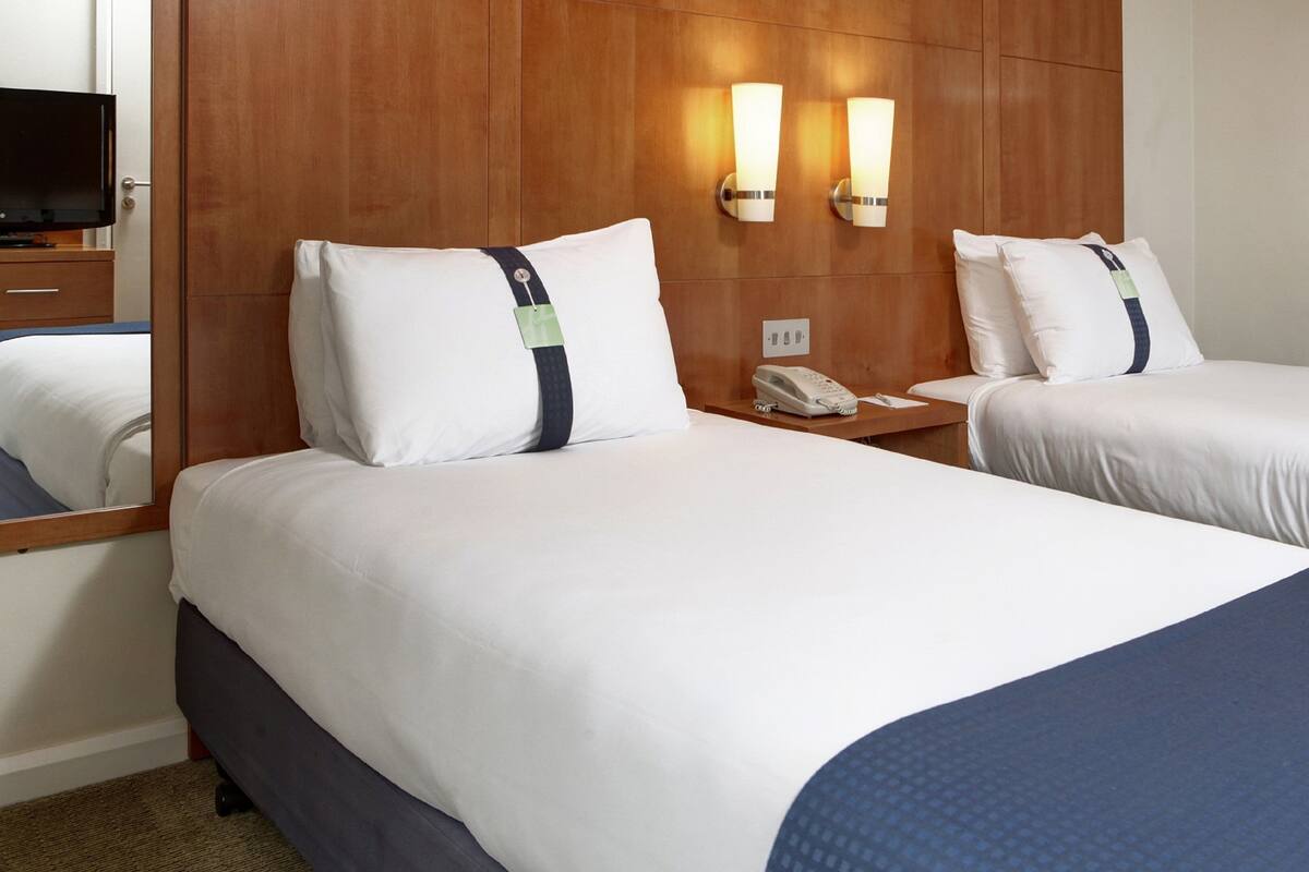Quarto de hotel com duas camas, cabeceira de madeira, luminárias laterais, telefone e espelho.