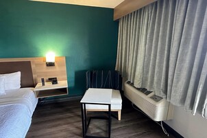 Quarto de hotel com cama, escrivaninha, cadeira, aquecedor fixado na parede e janela com cortinas.