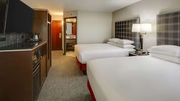 Quarto de hotel com uma cama grande, uma cama menor, uma escrivaninha com televisão de tela plana, uma luminária e um espelho.
