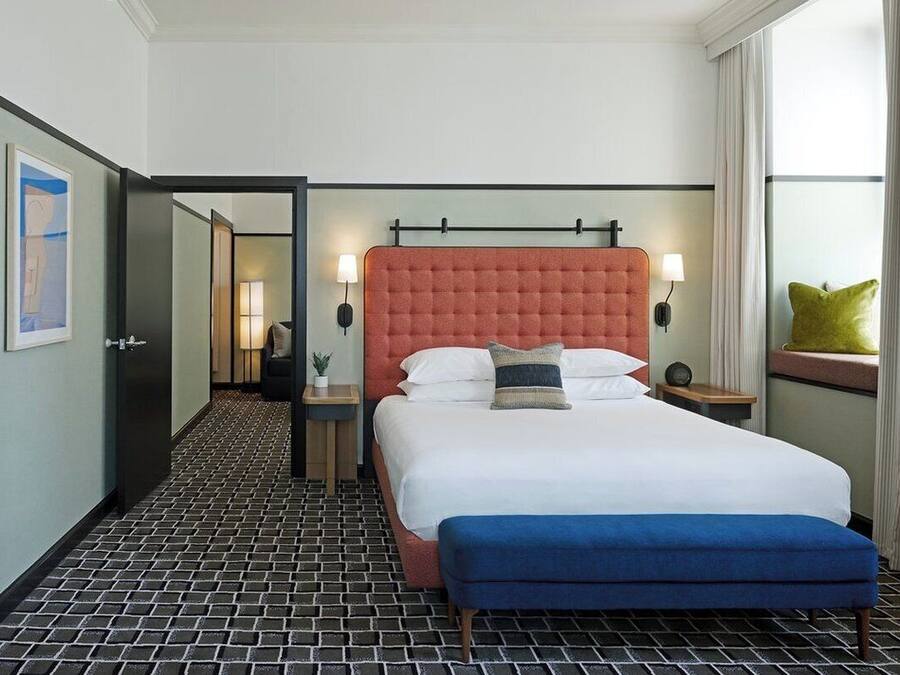 Quarto de hotel com uma cama grande, cabeceira, mesas de cabeceira, janela com cortinas e piso com padrão de tijolos.
