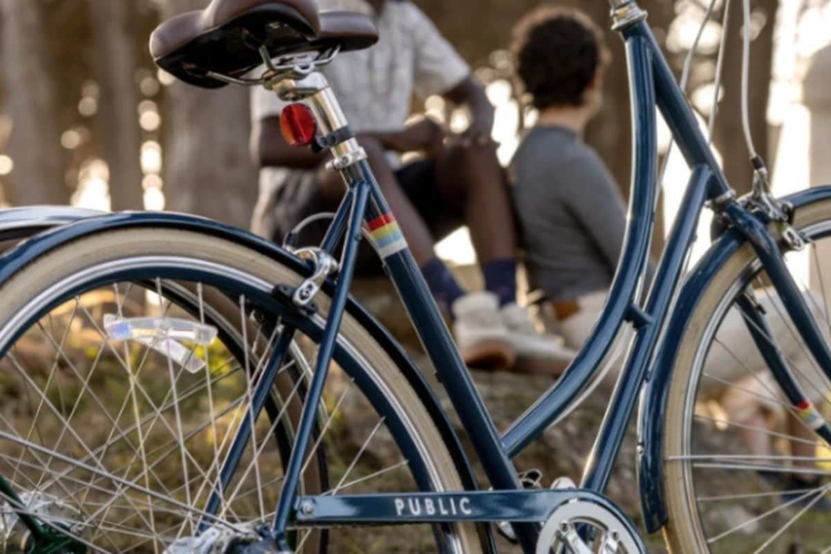 Uma bicicleta com a etiqueta 'PUBLIC' na estrutura, posicionada em um ambiente natural com árvores e pessoas ao fundo.