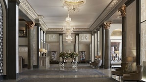 Um amplo saguão com lustre, colunas de mármore e uma mesa central decorada com flores.