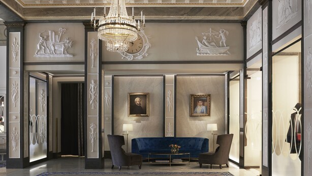 Um hall de entrada luxuoso com um lustre imponente, paredes de mármore e esculturas clássicas. Há duas poltronas, um sofá azul e uma mesa de centro com um vaso.