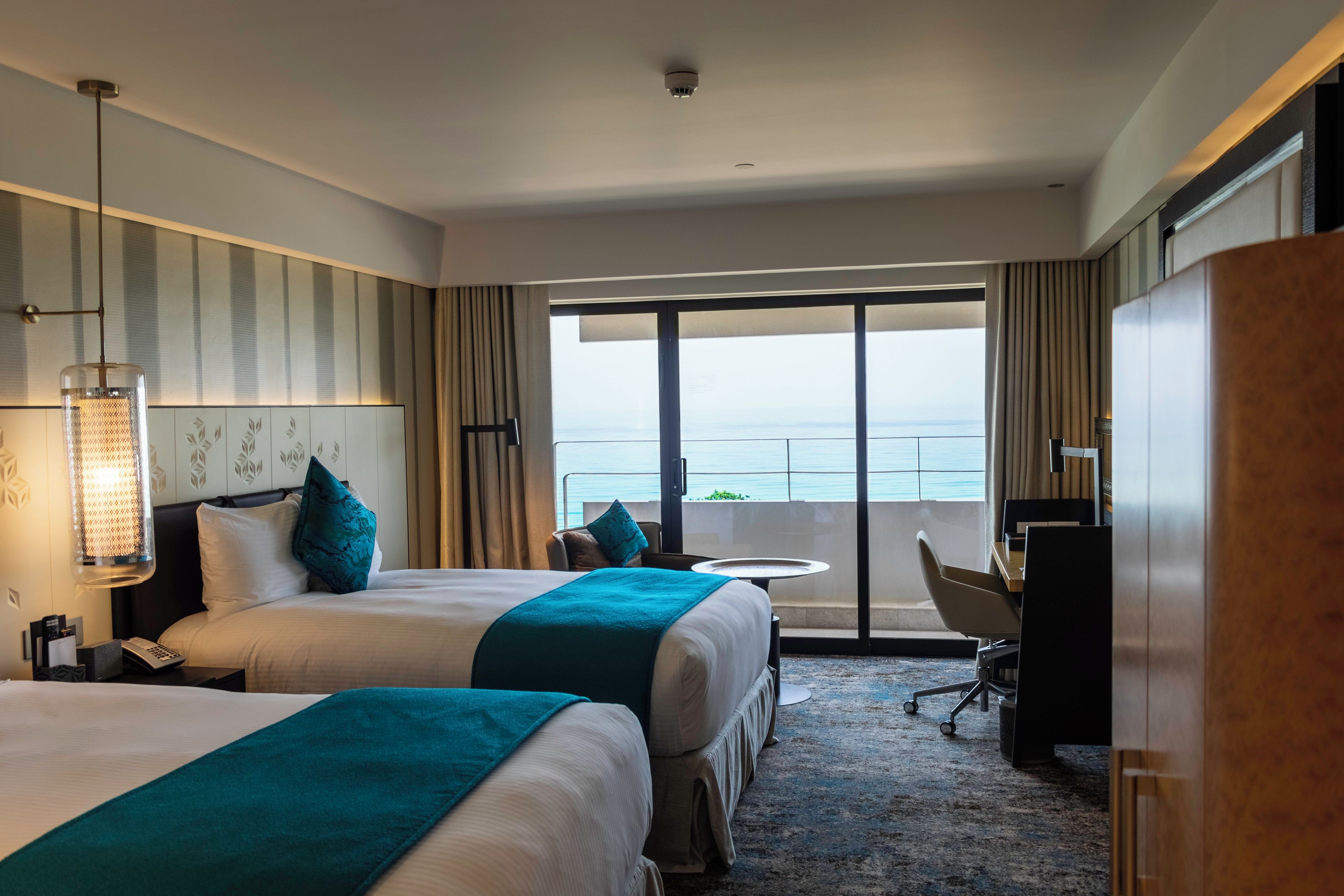 Quarto de hotel com duas camas, uma escrivaninha, uma cadeira, uma mesa pequena e vista para o mar através de uma janela grande.