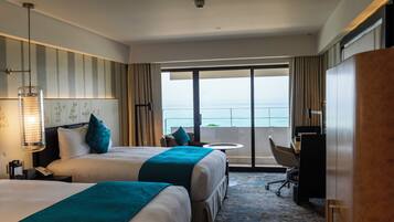 Quarto de hotel com duas camas, uma escrivaninha, uma cadeira, uma mesa pequena e vista para o mar através de uma janela grande.