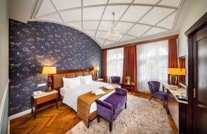 Quarto de hotel com uma cama grande, uma escrivaninha, uma cadeira, uma mesinha de cabeceira, um abajur e um painel decorativo.