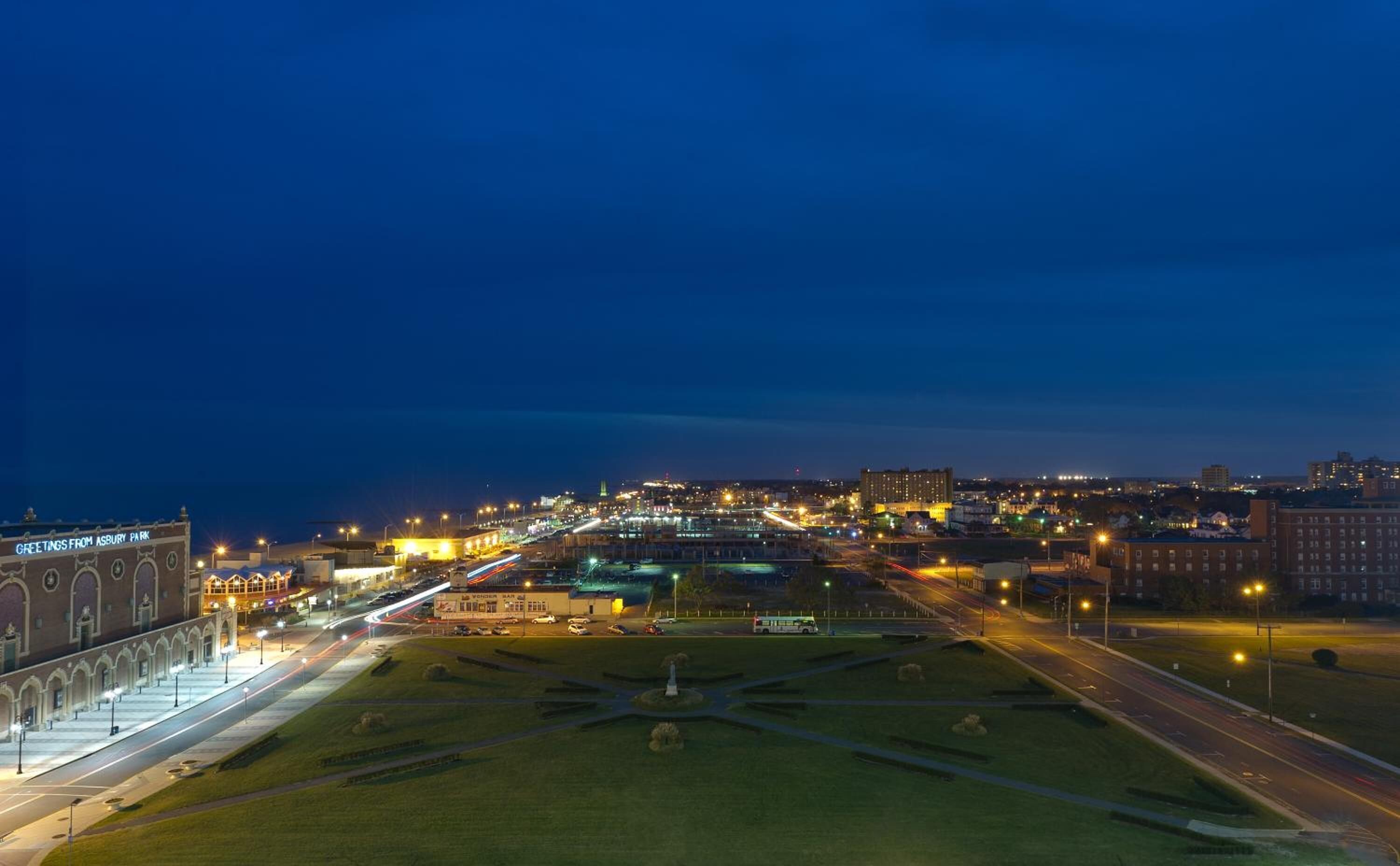 Um horizonte urbano noturno com um edifício em destaque, exibindo uma placa com a mensagem 'Greetings from Asbury Park'.