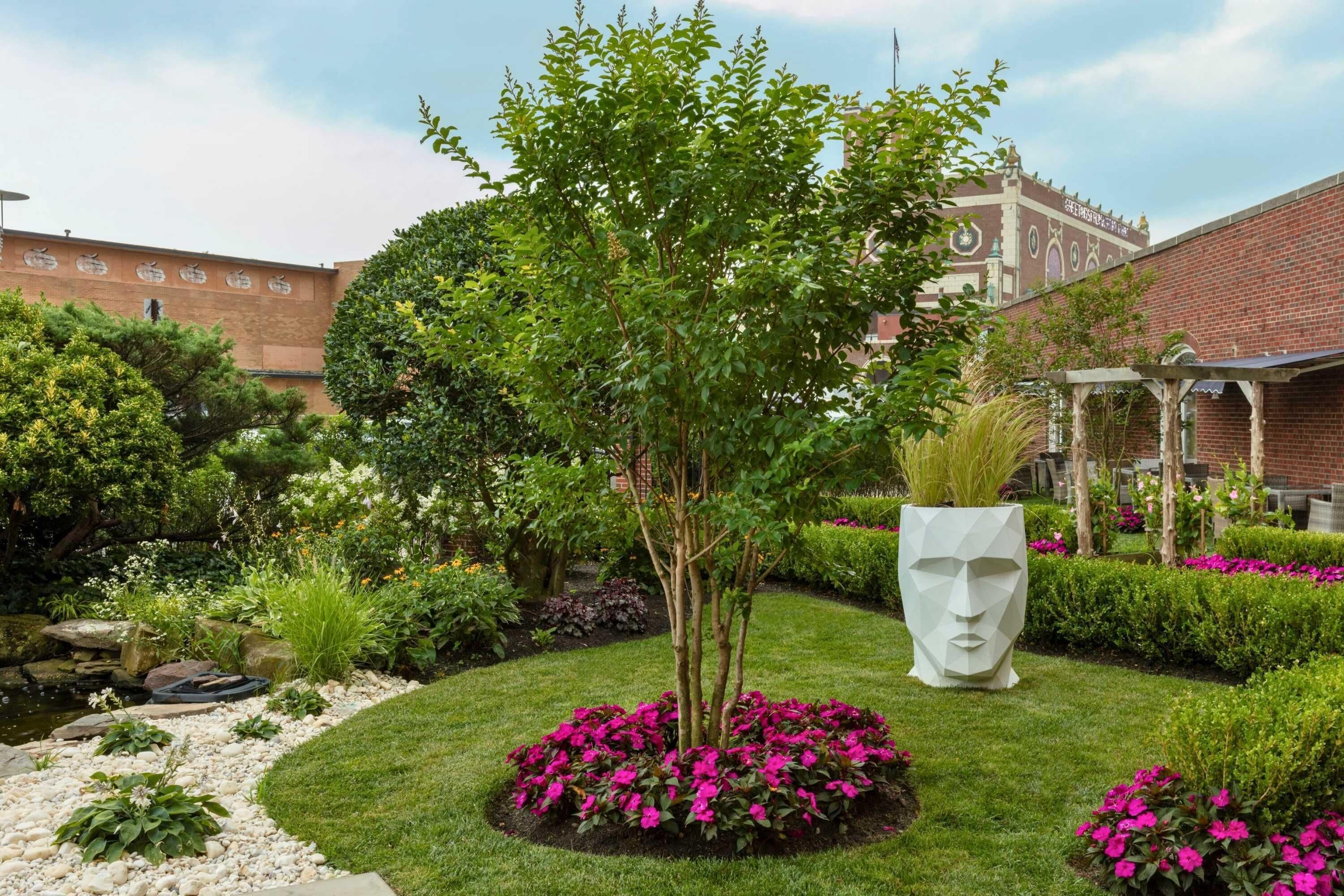 Um jardim bem cuidado com diversas plantas e flores, um elemento escultórico marcante e um edifício com fachada de tijolos ao fundo.