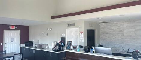 Uma área de recepção de hotel com balcão, mesa de jantar e cadeiras. Há uma placa indicando uma saída e um local para registro de hóspedes.