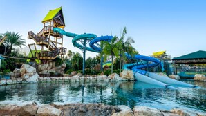 Um parque aquático com um grande escorregador de água azul, uma torre de observação de madeira e uma área de piscina rodeada por pedras.