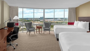 Quarto de hotel com uma cama grande, uma escrivaninha com computador, duas cadeiras, uma mesinha e vista para a cidade.