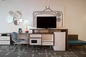 Quarto compacto com cama, escrivaninha, TV de tela plana, micro-ondas e geladeira.