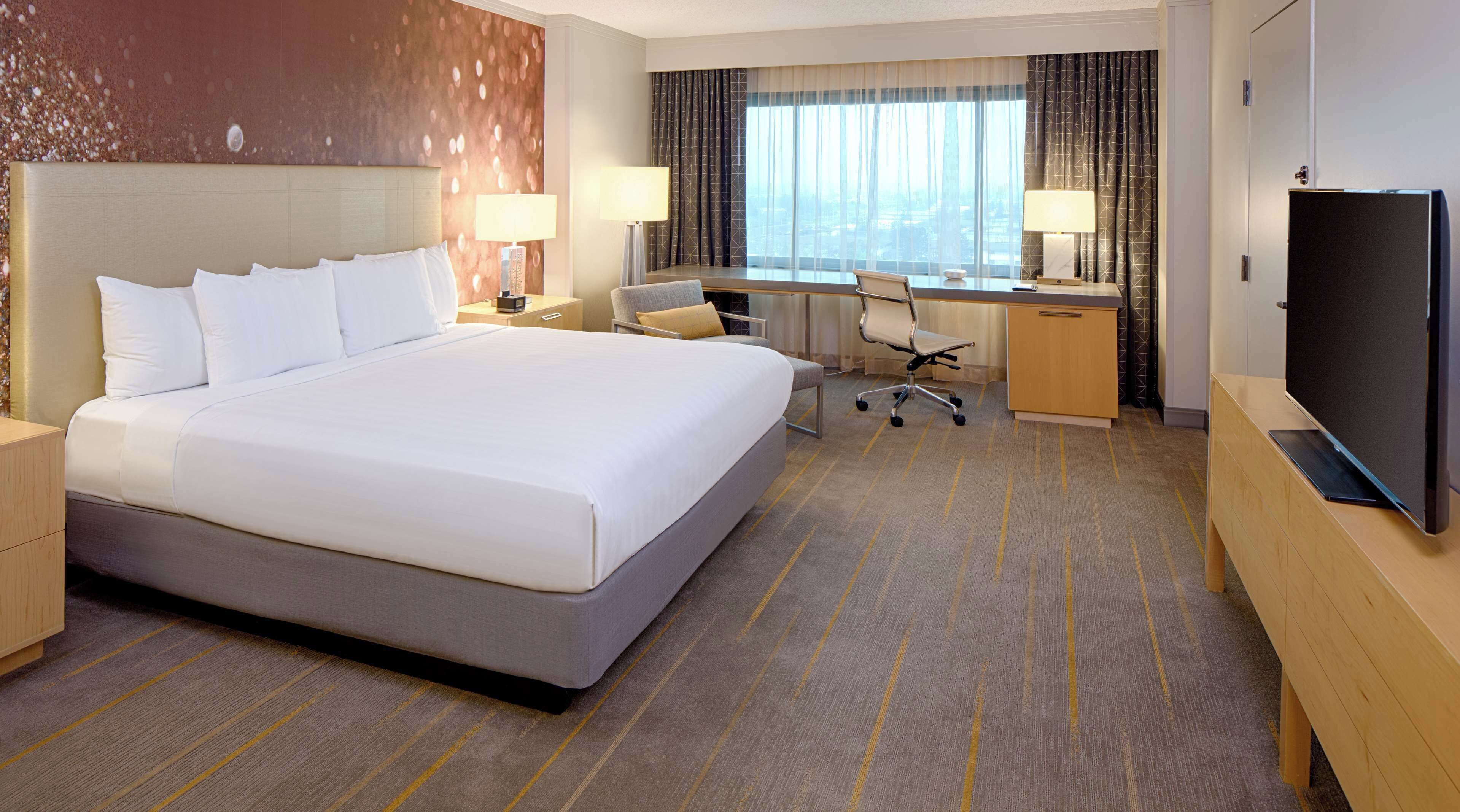 Quarto de hotel com uma cama grande, uma escrivaninha, uma cadeira, televisão e vista para a cidade.