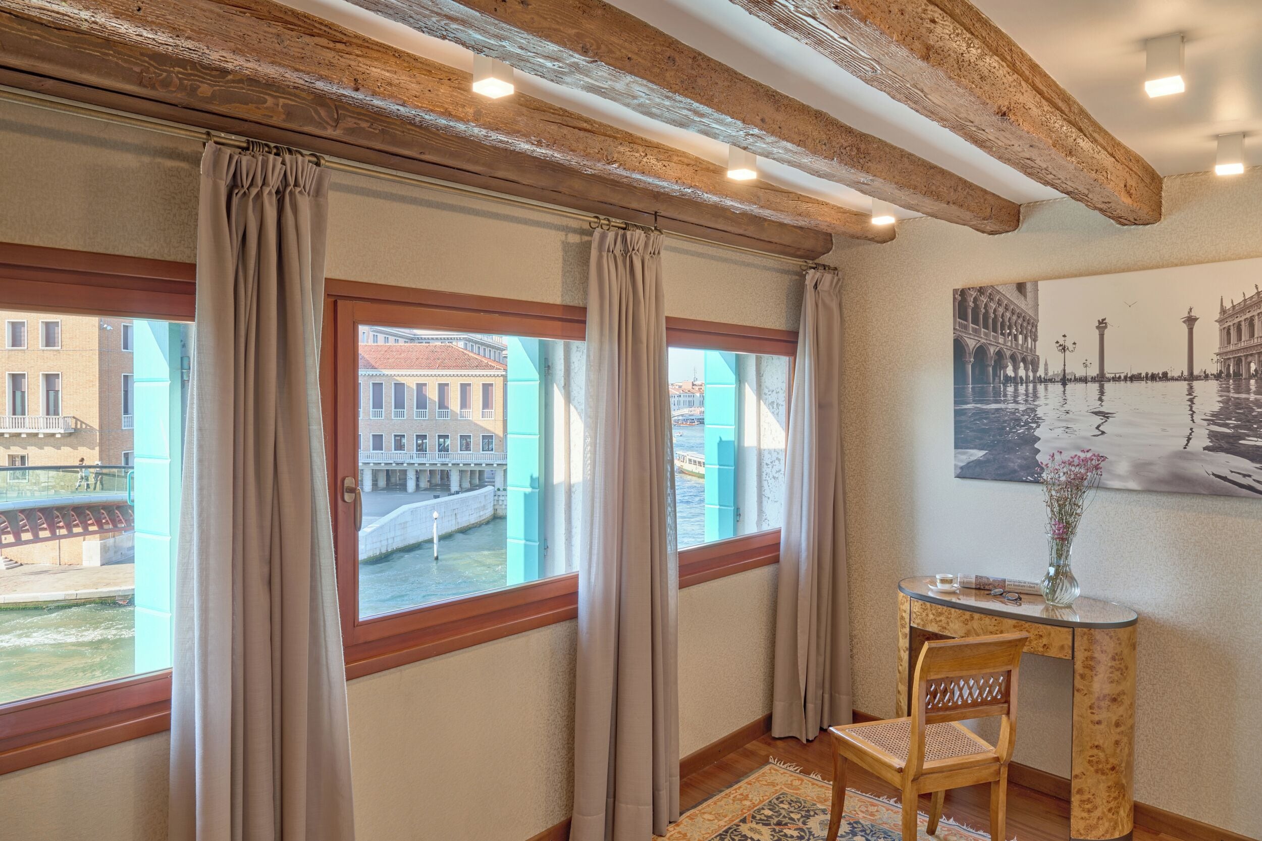 Quarto com uma janela grande, uma cadeira de madeira, uma mesa pequena com um vaso e um quadro de uma paisagem urbana.