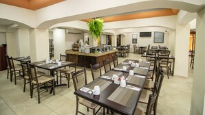 Uma sala de jantar espaçosa com várias mesas e cadeiras, uma ilha central com uma planta e uma área de cozinha ao fundo.