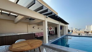 Área de piscina no terraço com mesa de madeira, cadeiras de vime branco, espaço de estar coberto e vista para a cidade.
