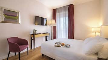 Quarto de hotel com cama, escrivaninha com computador, cadeira, televisão e janela com cortinas.