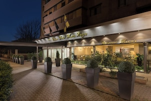 Entrada de hotel com fachada moderna, plantas em vasos grandes e área de acesso bem iluminada.