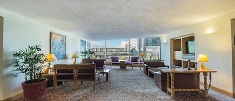 Um saguão de hotel com carpete estampado, área de estar com cadeiras e mesas, uma planta em vaso e uma grande janela com vista para edifícios.