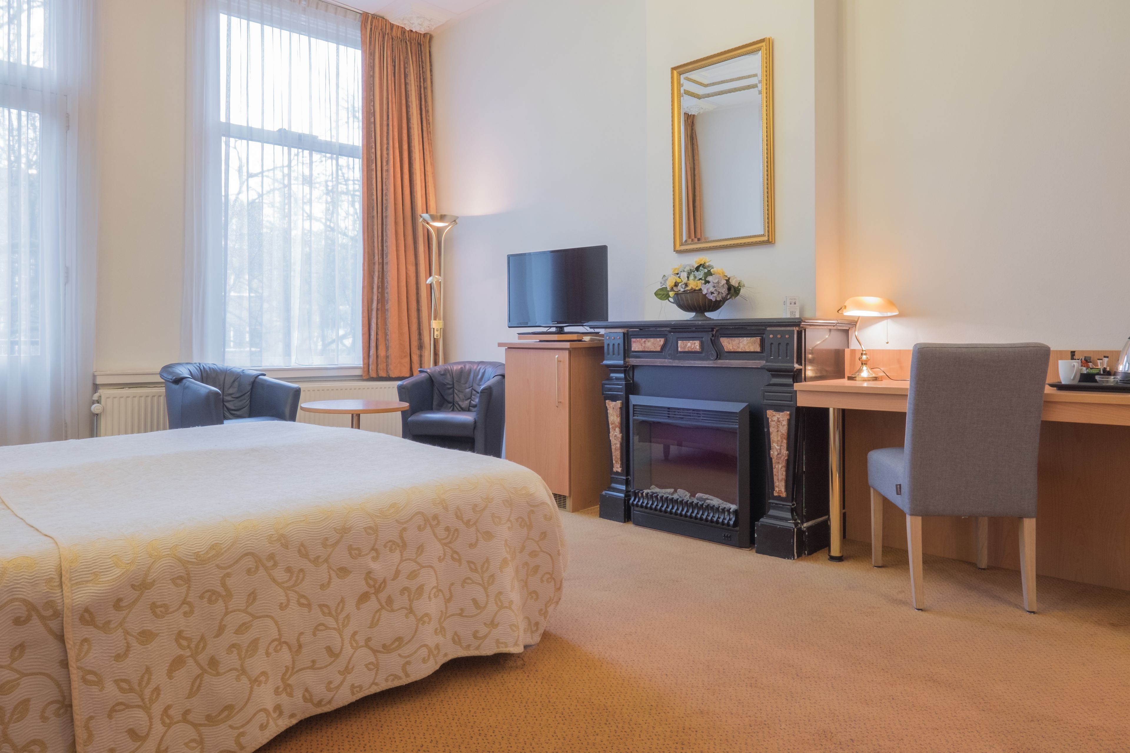 Quarto de hotel com cama, escrivaninha, cadeira, televisão, lareira e uma janela grande com cortinas.