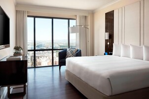 Quarto de hotel com uma cama grande, uma escrivaninha com televisão de tela plana, uma cadeira e vista da cidade através de amplas janelas.