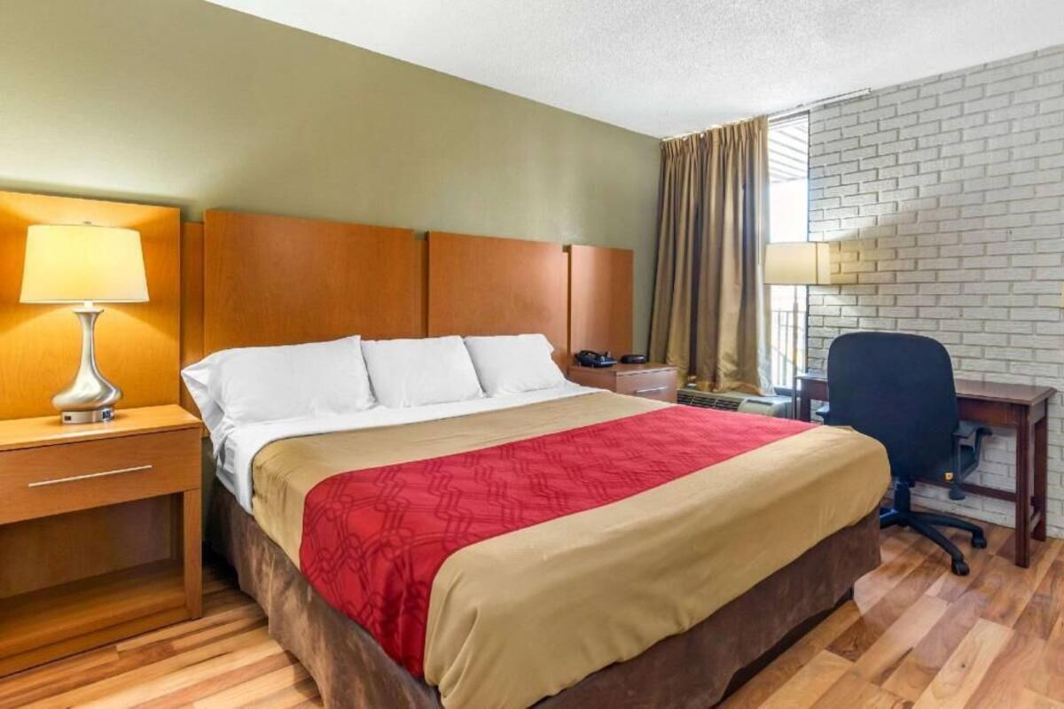 Quarto de hotel com cama, escrivaninha, cadeira, abajur e parede com padrão de tijolos.