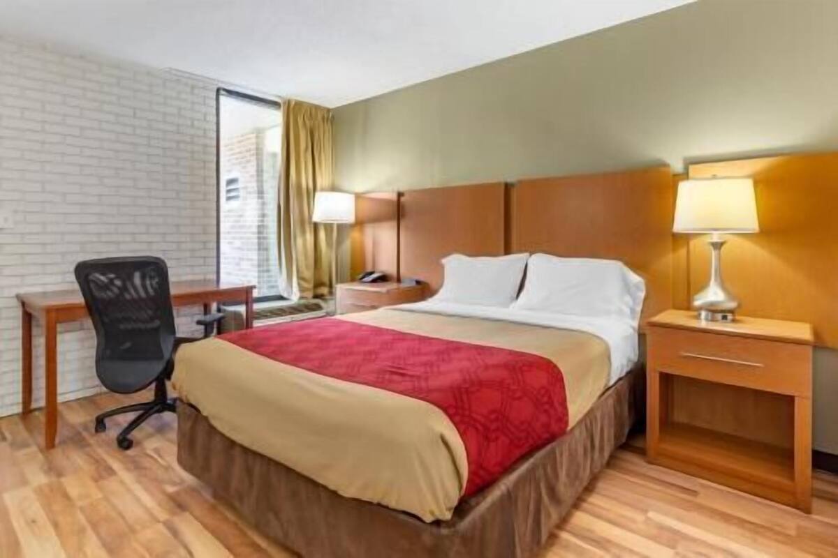 Quarto de hotel com cama, mesas de cabeceira, escrivaninha, cadeira e janela com cortinas.