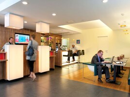 Uma área de recepção de hotel, com balcão de recepção, zona de bar com prateleiras de garrafas e espaço de estar com mesas e cadeiras.