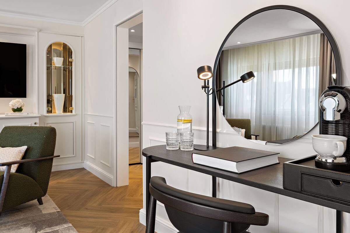 Quarto de hotel moderno com um espelho grande, uma máquina de café, uma escrivaninha com um livro e uma poltrona verde.