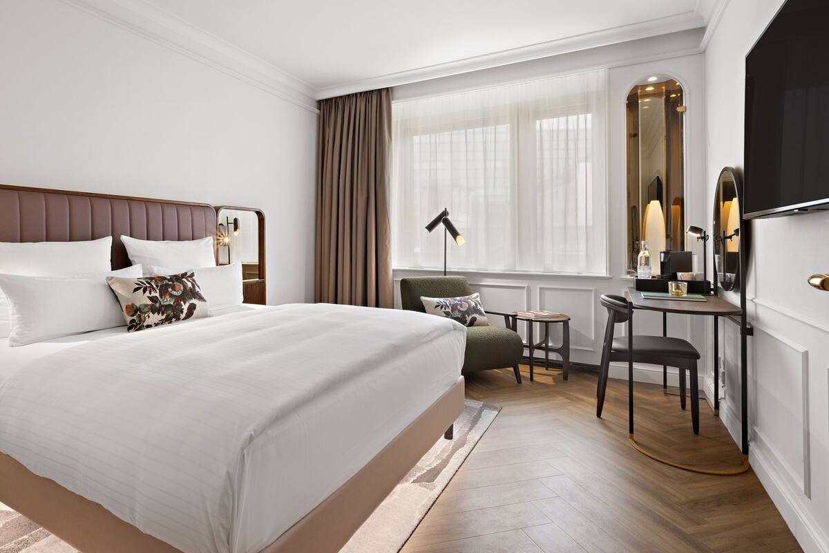 Quarto de hotel com uma cama grande, uma escrivaninha com cadeira, um espelho e uma janela com cortinas.