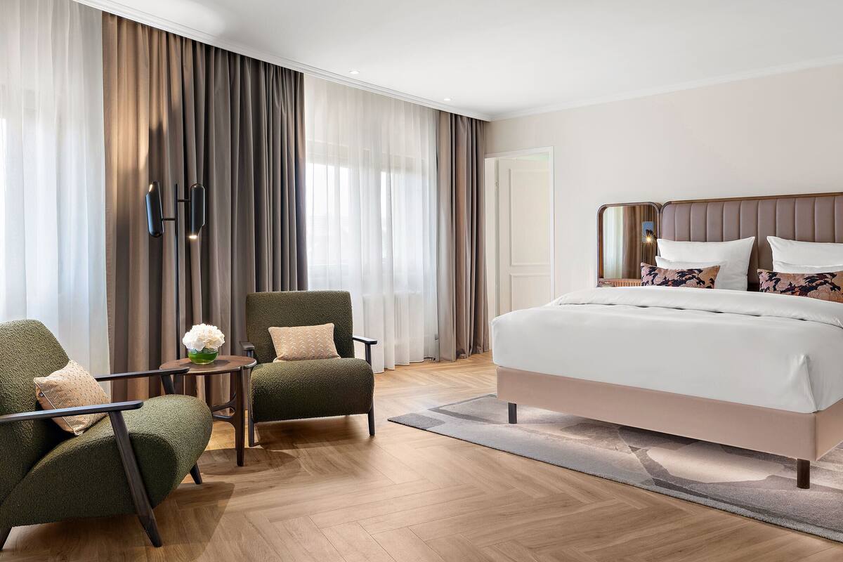 Quarto de hotel moderno com uma cama grande, duas poltronas, uma mesinha com um vaso de flores e uma janela ampla com cortinas.