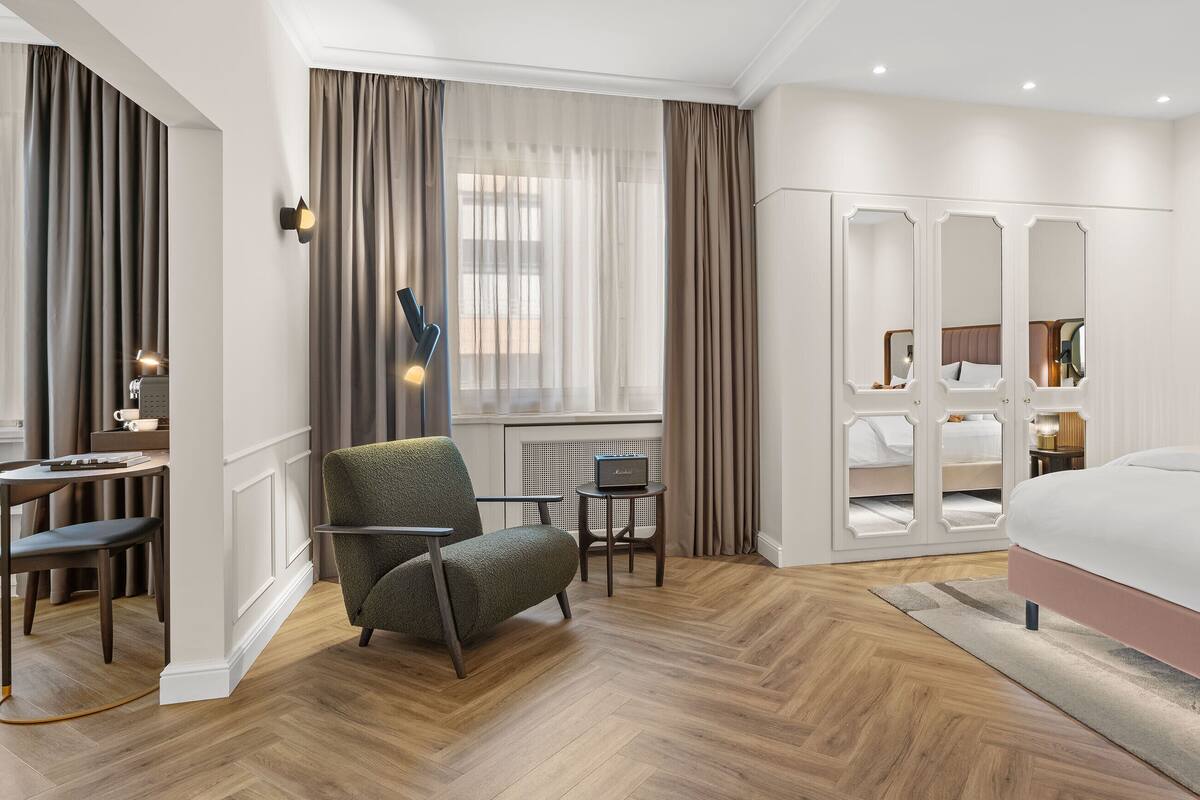 Quarto de hotel com uma cama, uma escrivaninha com cadeira, uma mesa pequena, uma poltrona, um espelho e uma janela com cortinas.