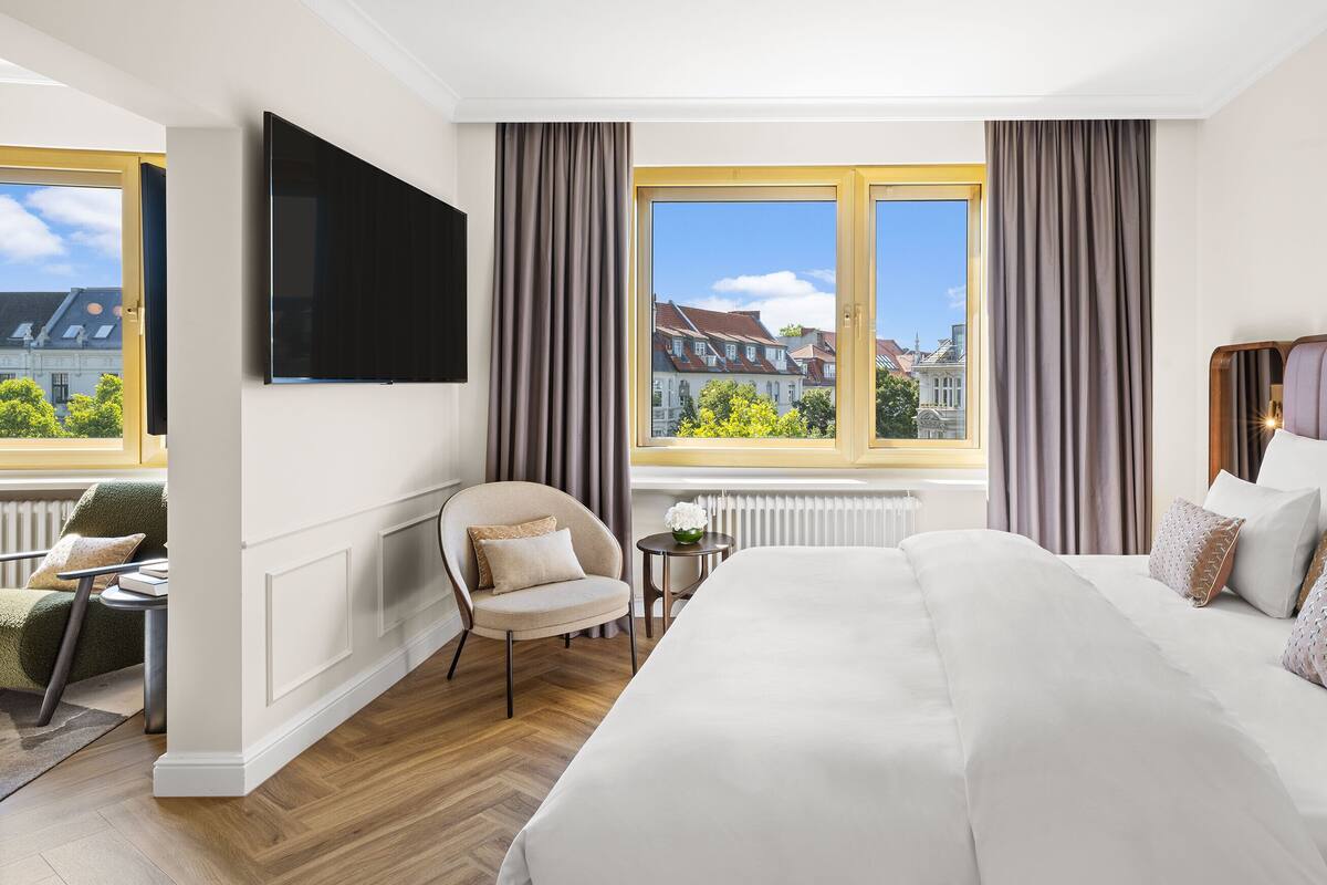 Quarto de hotel com uma cama grande, uma cadeira, uma mesa pequena e uma televisão fixada na parede.