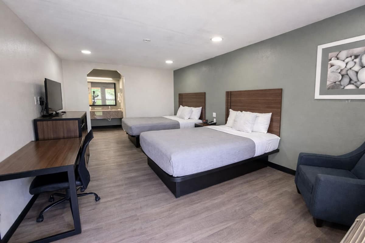 Quarto de hotel com duas camas, uma escrivaninha, uma cadeira, televisão e banheiro ao fundo.