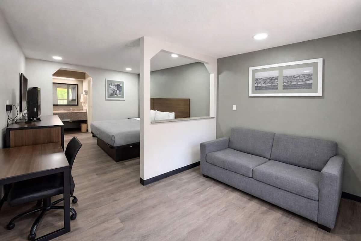 Quarto de hotel moderno com sofá cinza, secretária de madeira, cama com cabeceira branca e um quadro emoldurado na parede.