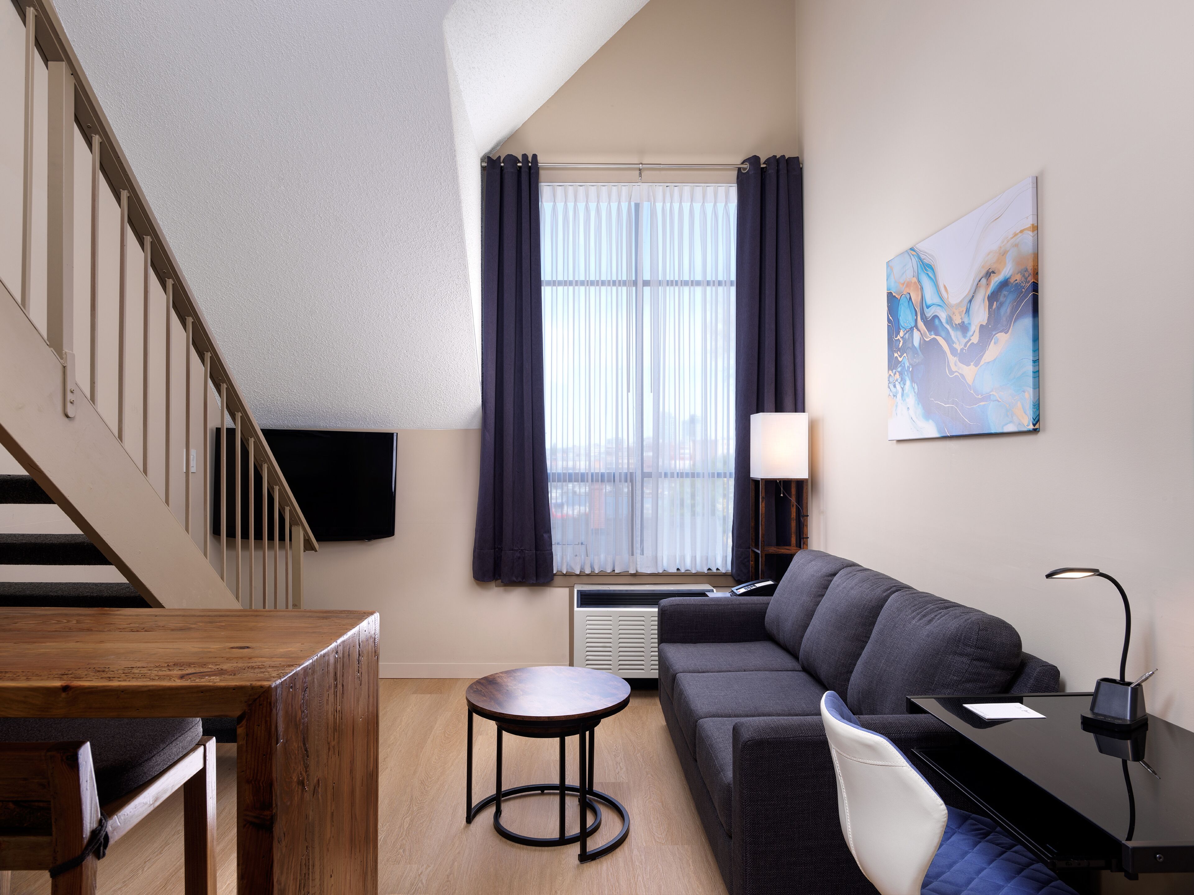 Uma sala de estar moderna com uma mesa de jantar de madeira, um sofá cinza, uma pequena mesa redonda, uma TV na parede e um quadro.