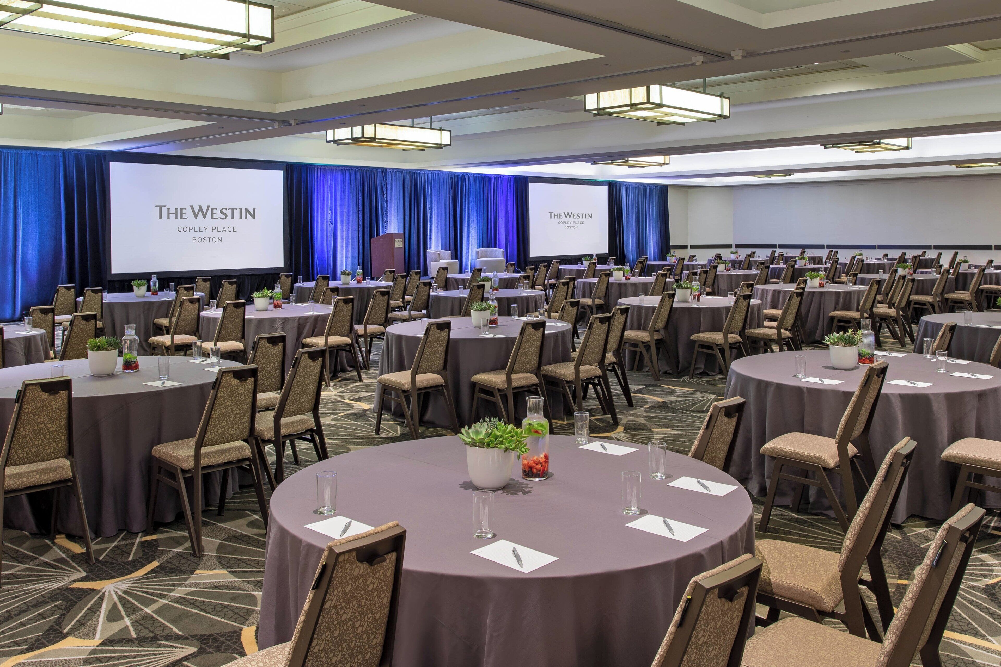 Uma sala de conferências no The Westin Copley Place, Boston, com várias mesas redondas, cadeiras e uma tela grande exibindo o nome do hotel.