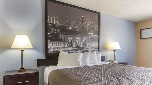 Quarto de hotel com uma cama grande, duas mesas de cabeceira com abajures, um quadro com pintura de paisagem urbana e uma janela com persianas.