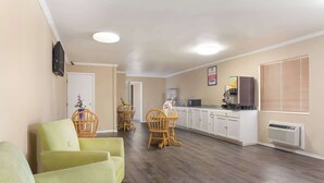 Uma sala de estar com sofá verde, uma área de jantar com cadeiras de madeira, uma cozinha com armários brancos e uma TV fixada na parede.