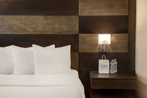 Quarto de hotel com cama arrumada, mesa de cabeceira com abajur e garrafa de água sobre ela.