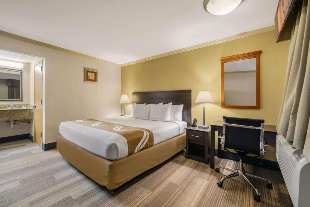 Quarto de hotel com cama, mesa de cabeceira, luminária e escrivaninha com cadeira.