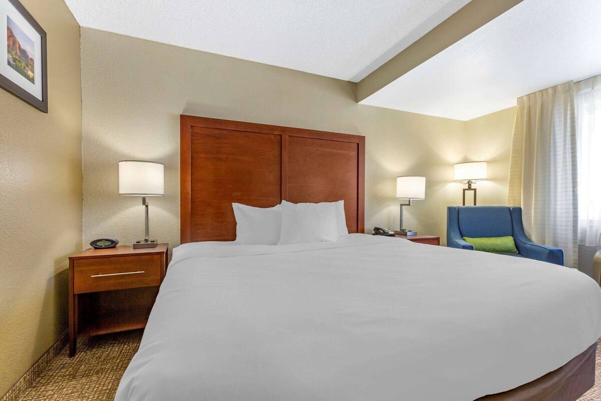 Quarto de hotel com uma cama grande, duas mesas de cabeceira, uma cadeira e uma luminária.